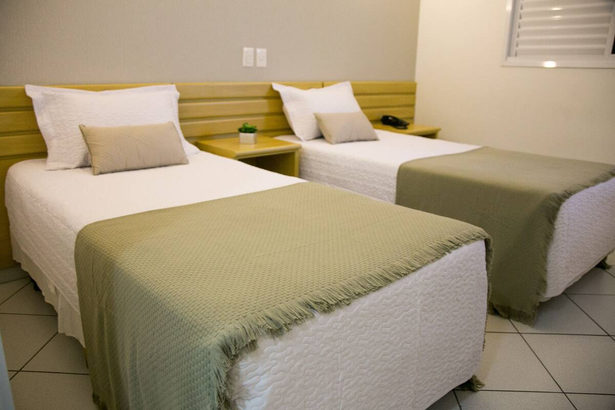 Duas camas de solteiro com roupa de cama branca e verde, uma cabeceira de madeira, uma pequena mesa de cabeceira com uma planta e uma tomada na parede.