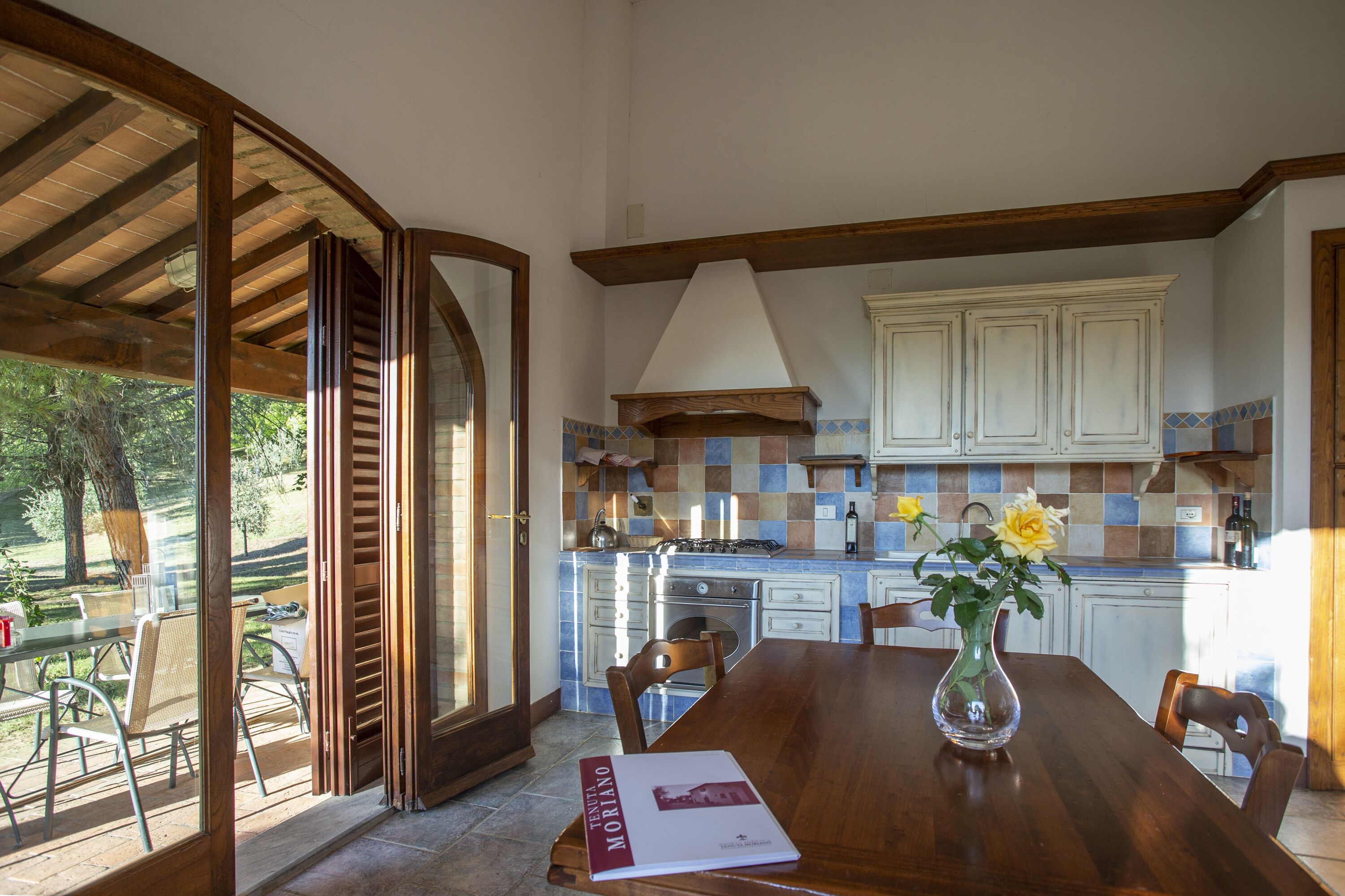Uma área de refeições com mesa de madeira, cadeiras e um vaso com flores. Há um livro sobre a mesa e uma vista da cozinha ao fundo.