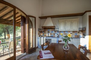 Uma área de refeições com mesa de madeira, cadeiras e um vaso com flores. Há um livro sobre a mesa e uma vista da cozinha ao fundo.