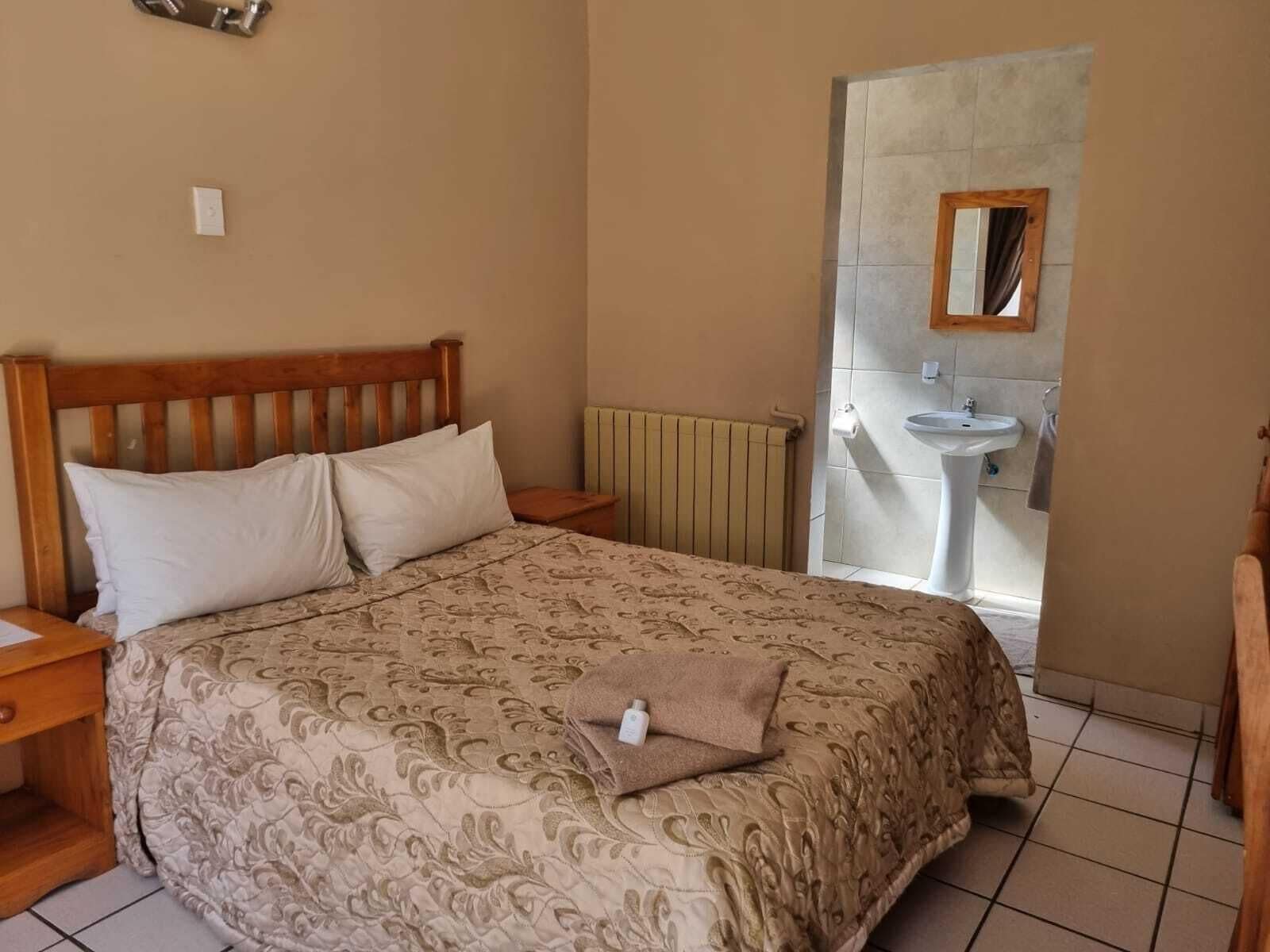 Quarto com cabeceira de madeira, colcha estampada, mesa de cabeceira com abajur e banheiro visível por uma porta aberta.