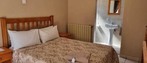 Quarto com cabeceira de madeira, colcha estampada, mesa de cabeceira com abajur e banheiro visível por uma porta aberta.
