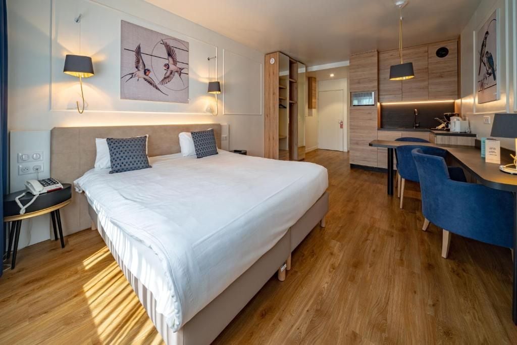 Quarto de hotel moderno com uma cama grande, mesas de cabeceira, escrivaninha e cozinha compacta.