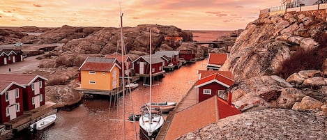 Uma vila costeira com casas tradicionais de telhados vermelhos, um litoral rochoso e barcos atracados na água.