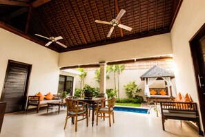 Uma área externa coberta com piscina, móveis de madeira, ventiladores de teto e uma estrutura com telhado de palha.
