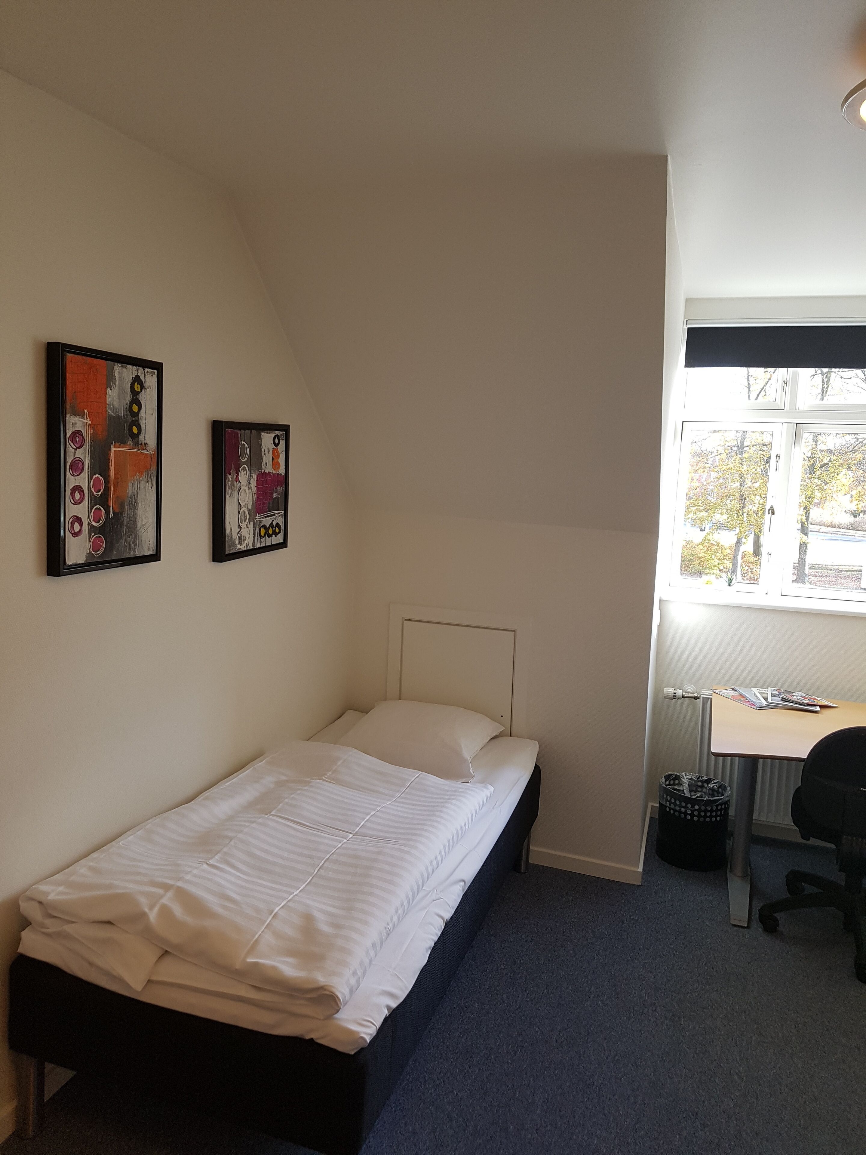 Um quarto pequeno e bem organizado, com uma cama de solteiro, uma escrivaninha e duas obras de arte emolduradas na parede.