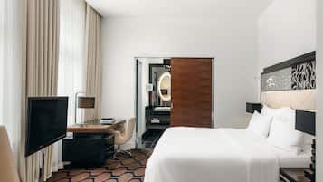 Quarto de hotel com cama, escrivaninha, televisão e banheiro visível através de uma porta aberta.