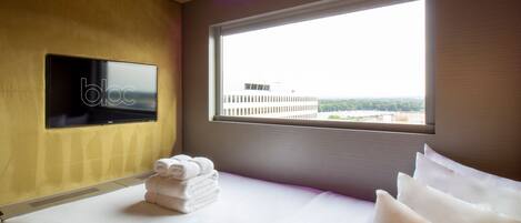 Quarto de hotel com cama, televisão fixada na parede, janela com vista para edifícios e uma pilha de toalhas.