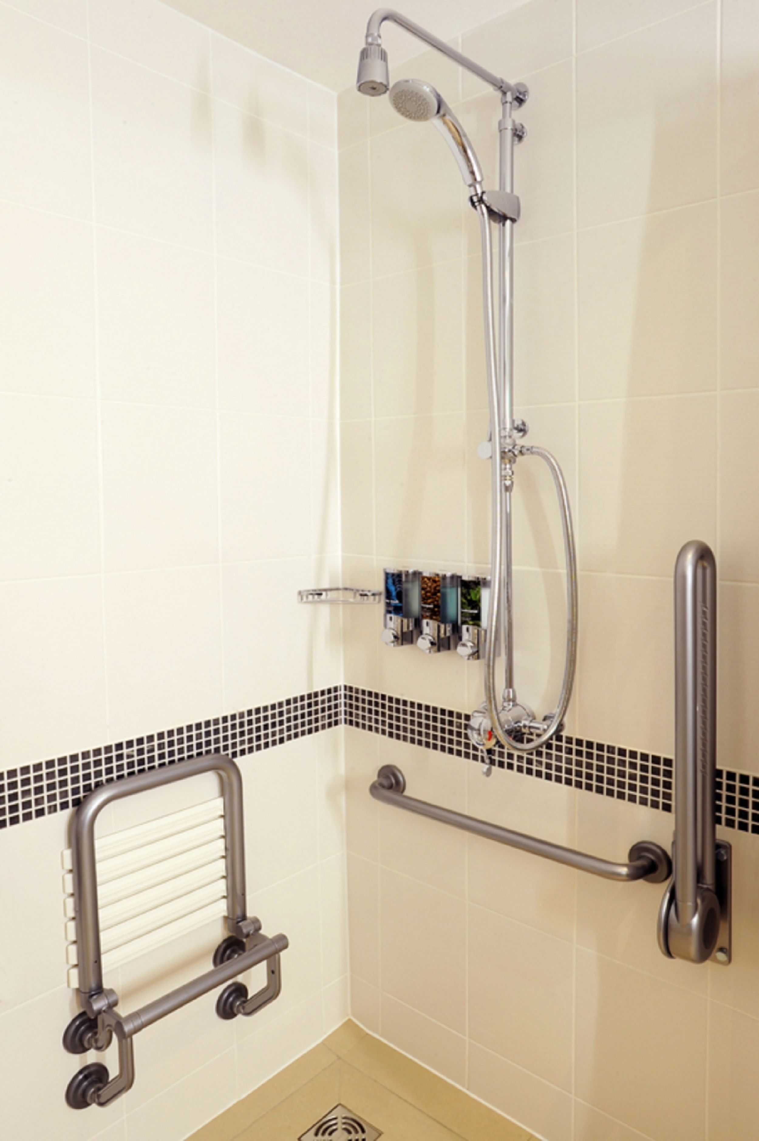 Área de chuveiro com chuveiro manual, chuveiro de chuva vertical e acessórios de chuveiro manual. Há uma barra de apoio instalada na parede e um porta-toalhas com uma toalha.