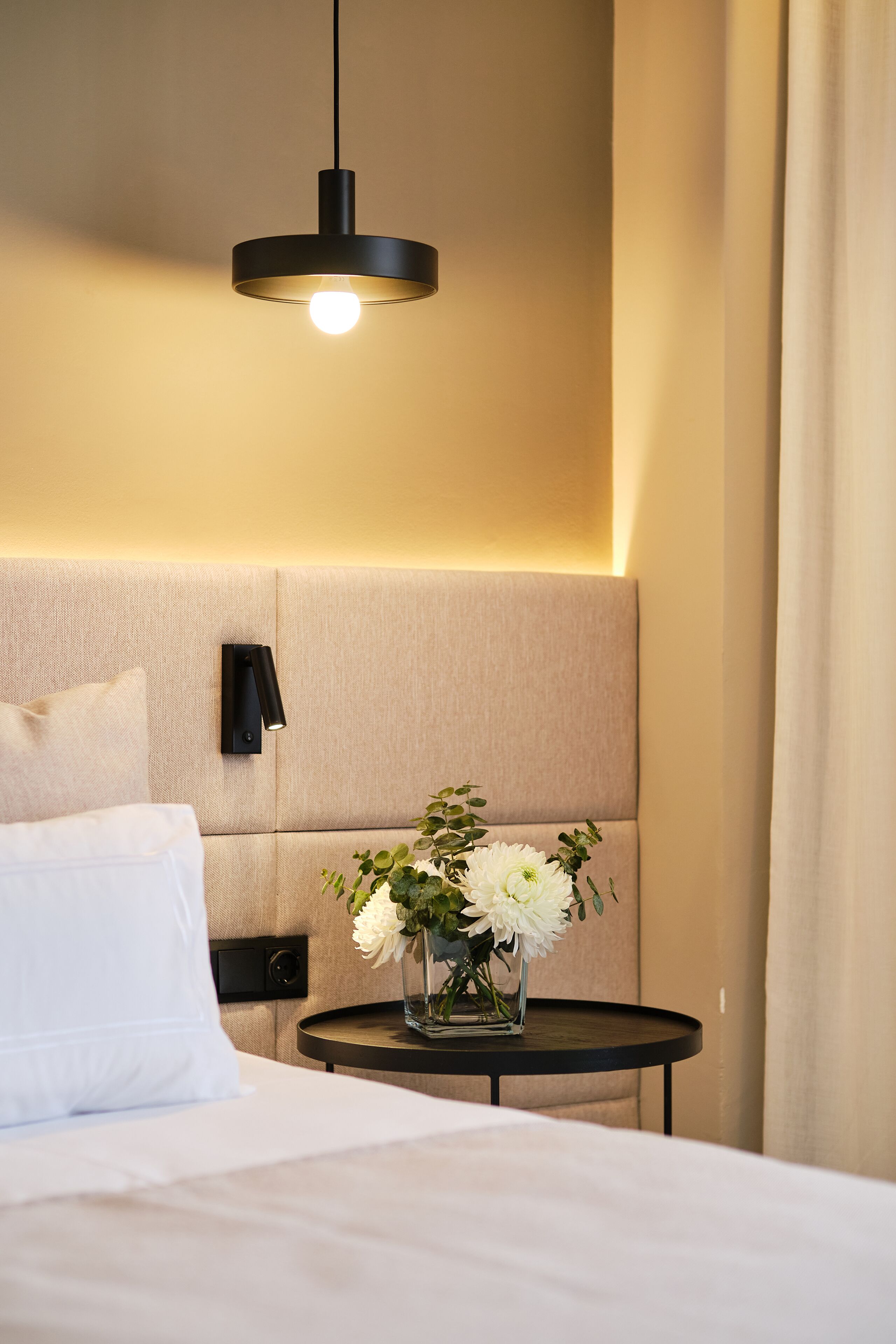Quarto de hotel com uma cama, uma mesa de cabeceira com um vaso de flores, uma luminária na parede e um painel de parede.