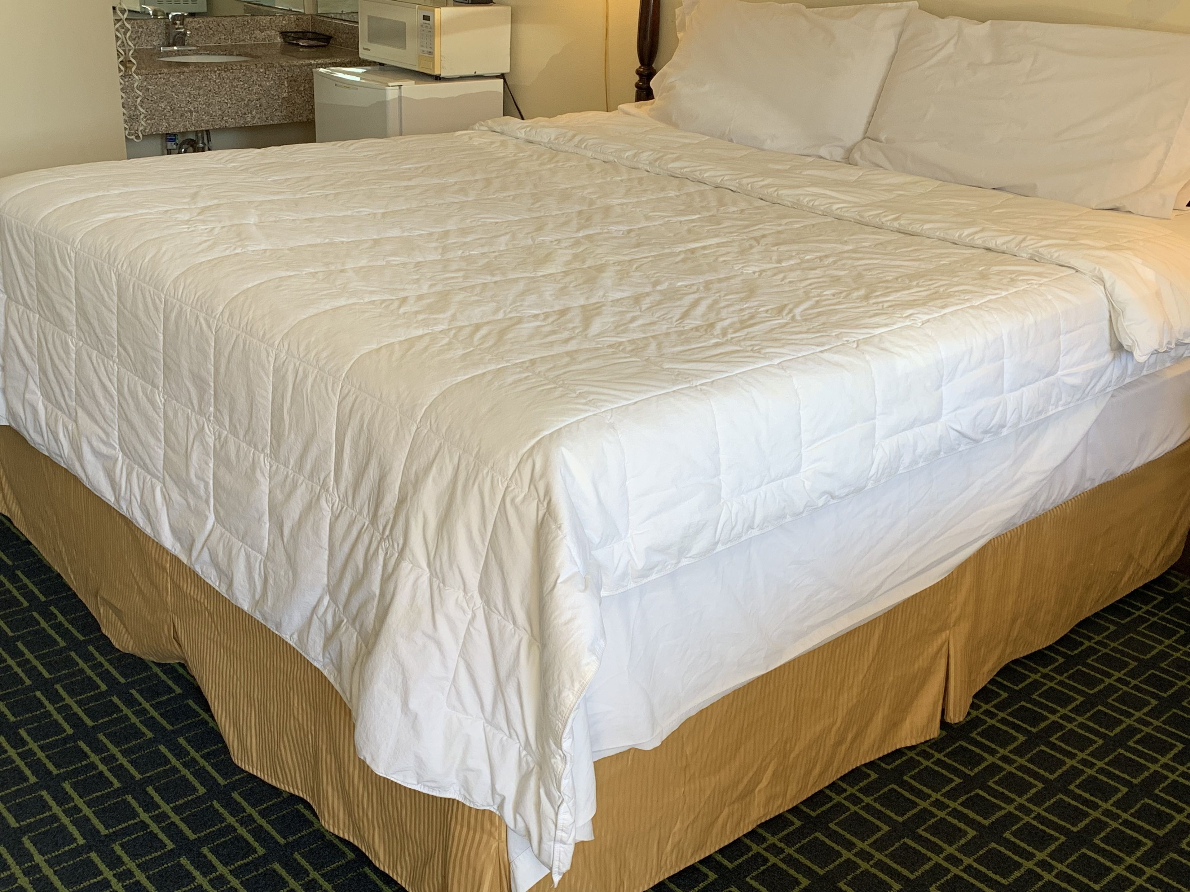Cama bem arrumada com colcha branca, cabeceira de madeira e um pequeno eletrodoméstico branco na mesa de cabeceira.