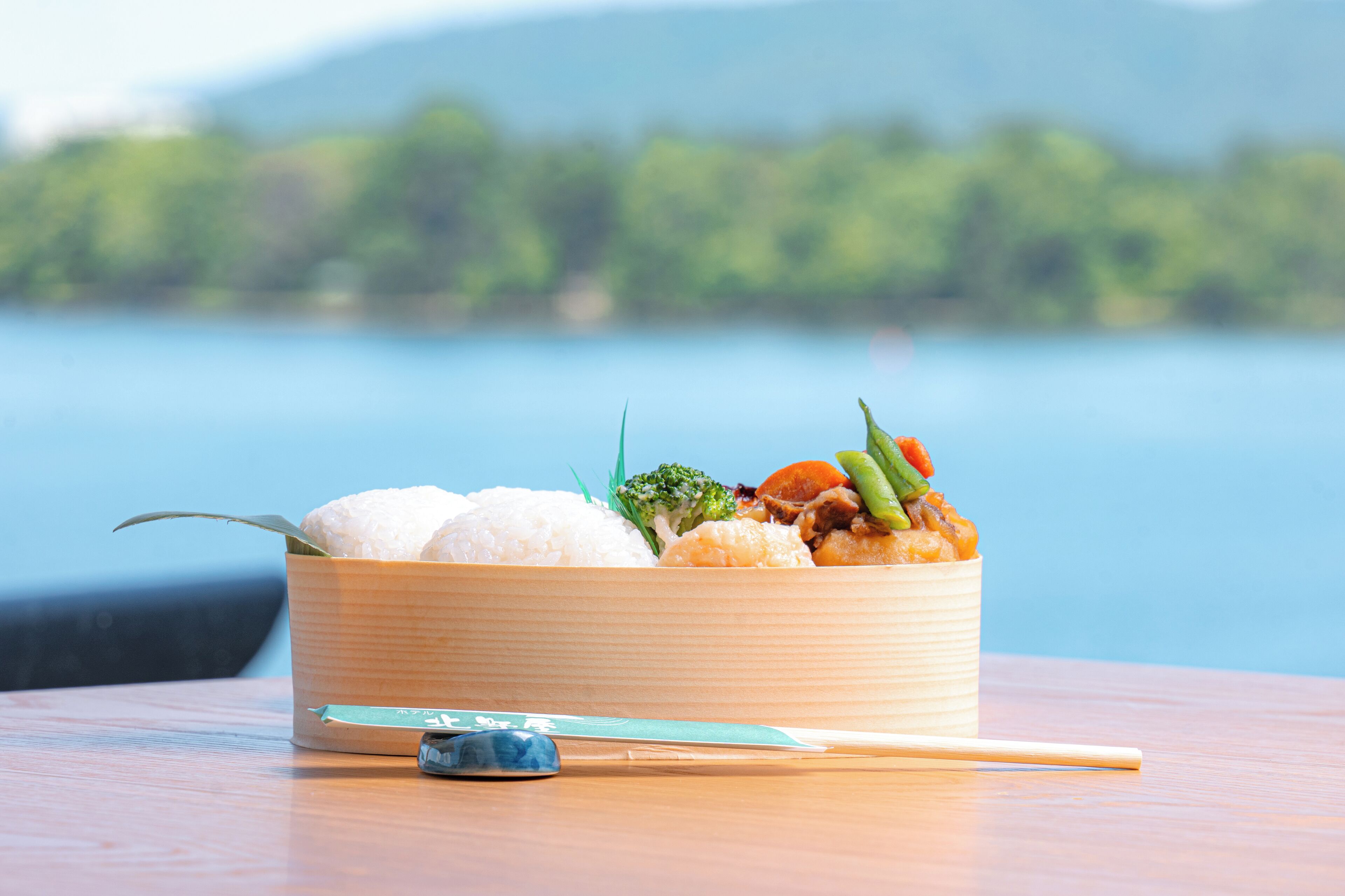 Uma mesa de madeira com uma tigela de bambu contendo arroz, legumes e um pedaço de carne, com um pauzinho de madeira e um objeto pequeno azul ao lado.