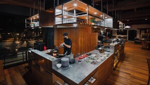 Uma cozinha moderna com um chef preparando comida, painéis de madeira e um espaço de bar com diversos pratos e utensílios.