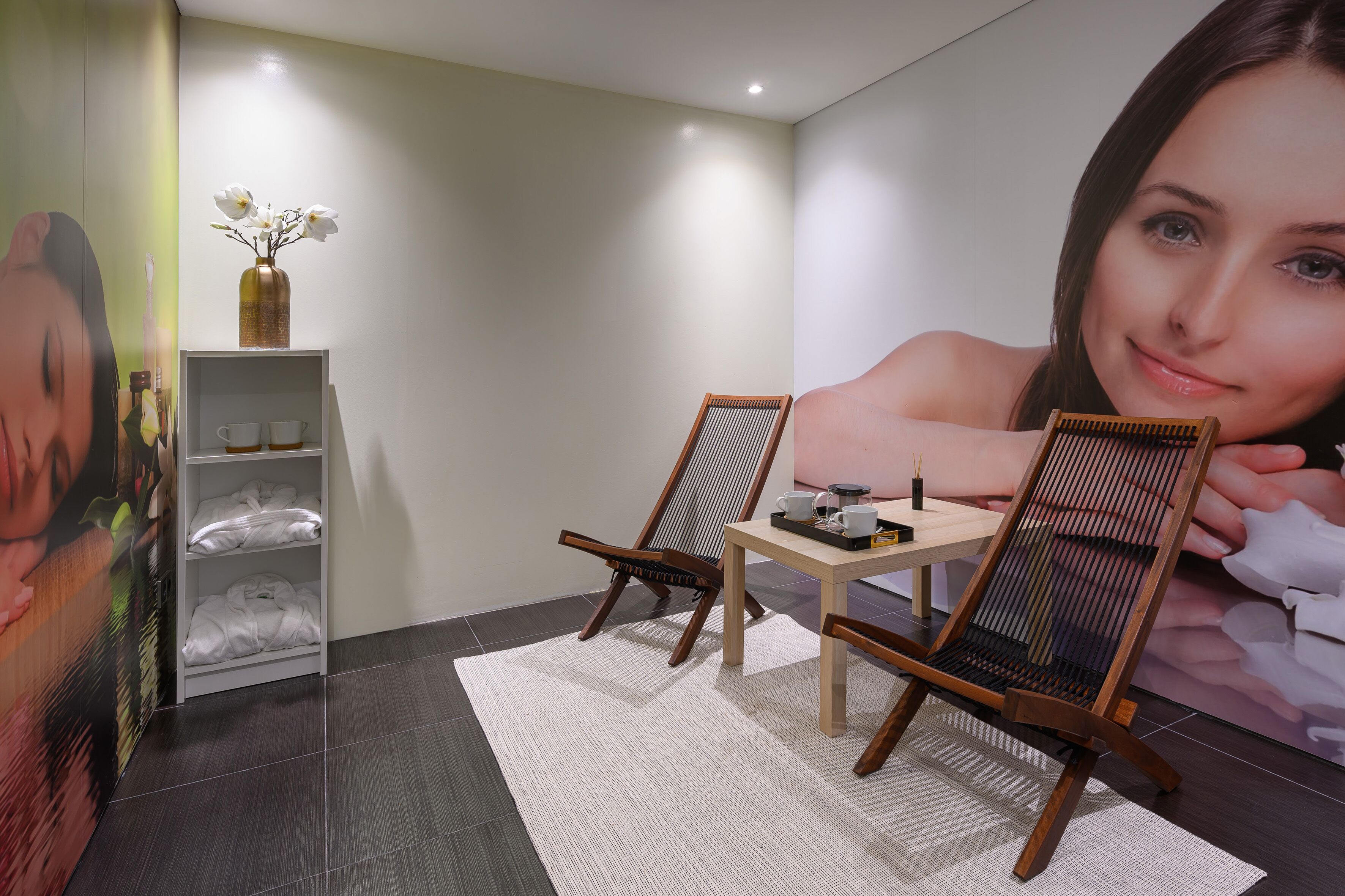 Um quarto com uma mesa de madeira, duas cadeiras de vime, uma prateleira branca com toalhas dobradas e um vaso com flores.