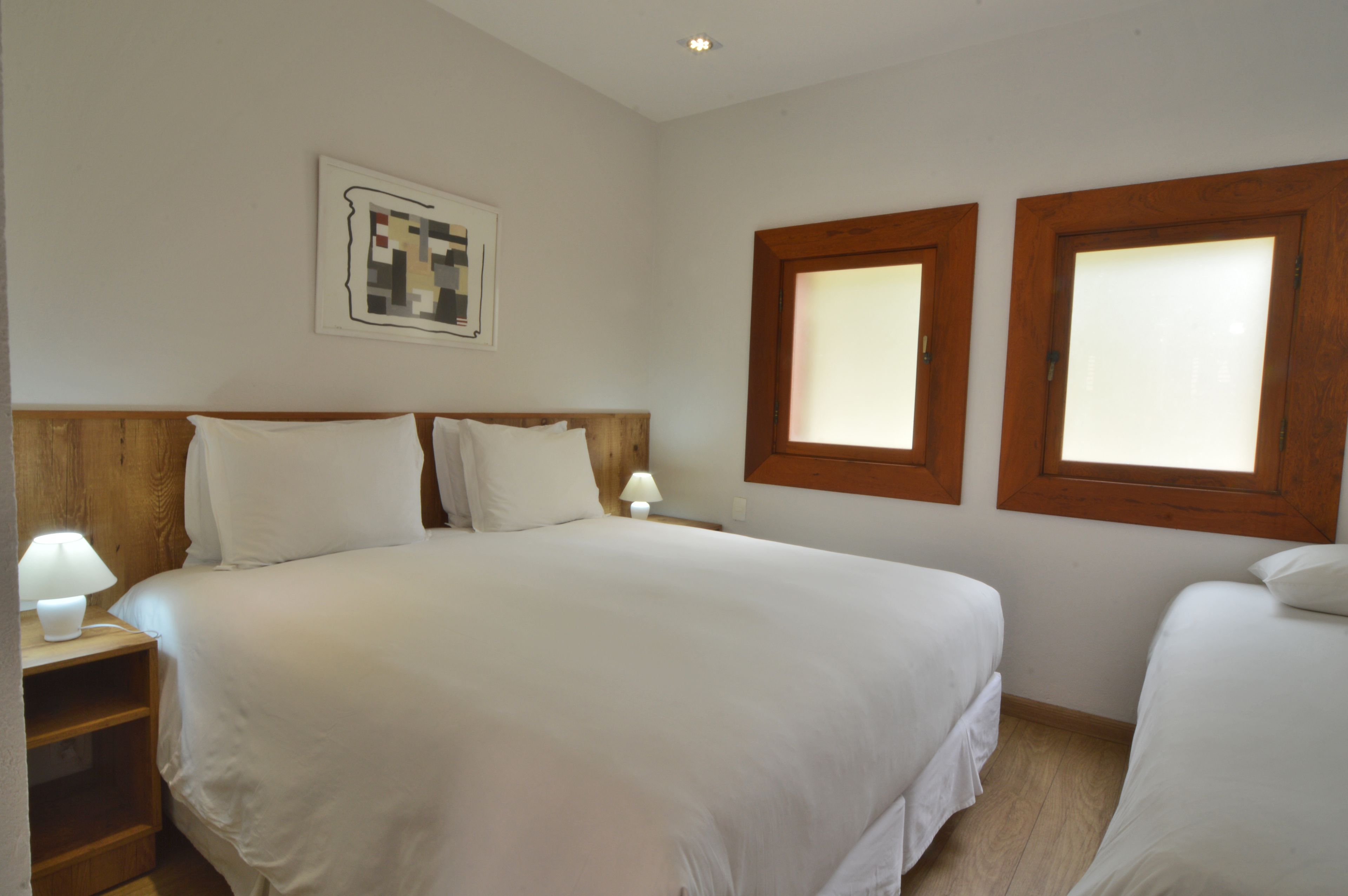 Quarto de hotel com uma cama grande, duas mesas de cabeceira com abajures e duas janelas com molduras de madeira.