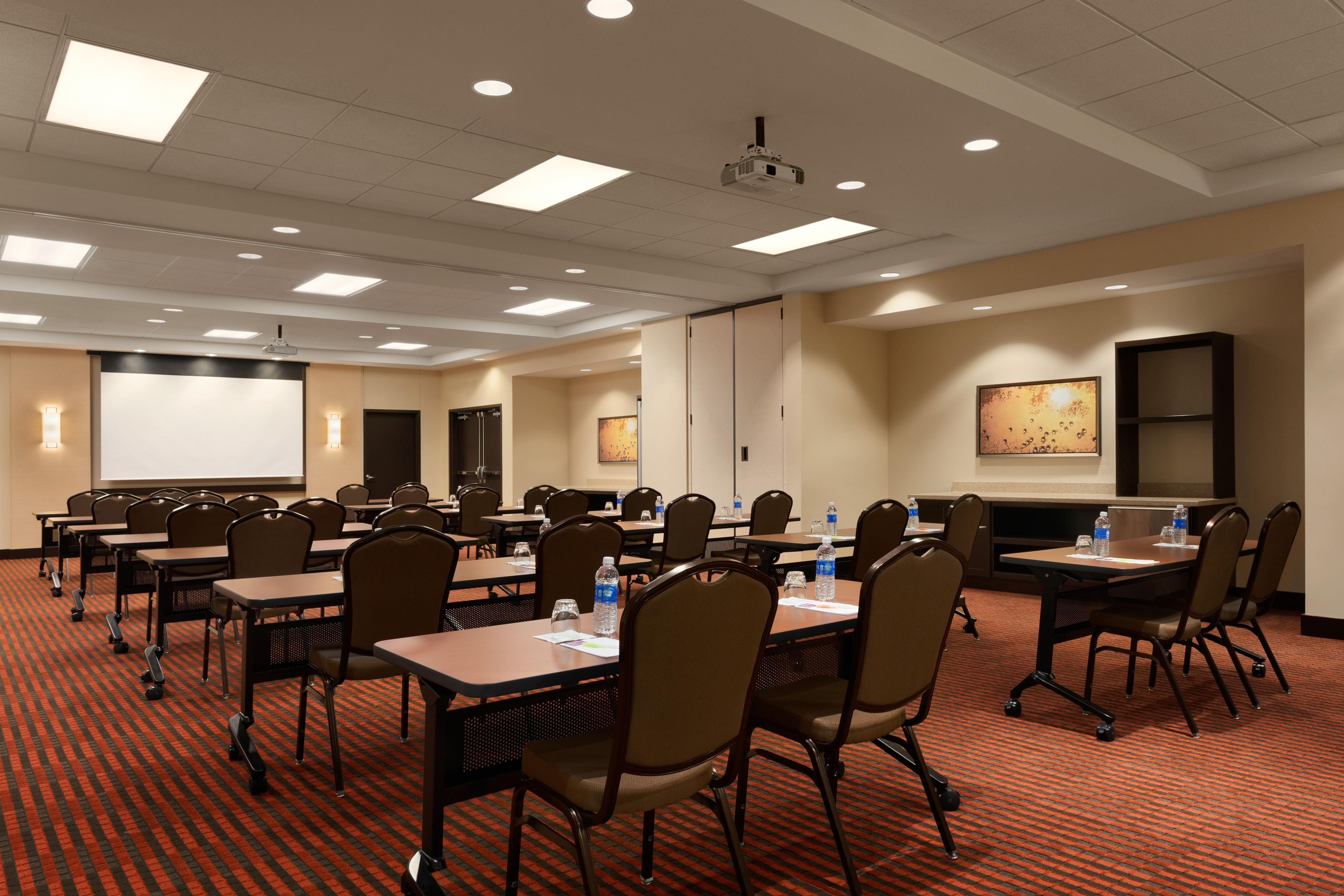 Sala de conferências com mesas, cadeiras, projetor e tela.