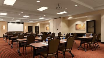 Sala de conferências com mesas, cadeiras, projetor e tela.