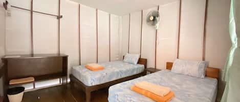 Quarto de hotel com duas camas, uma mesinha, um cesto de lixo e um ventilador de teto.