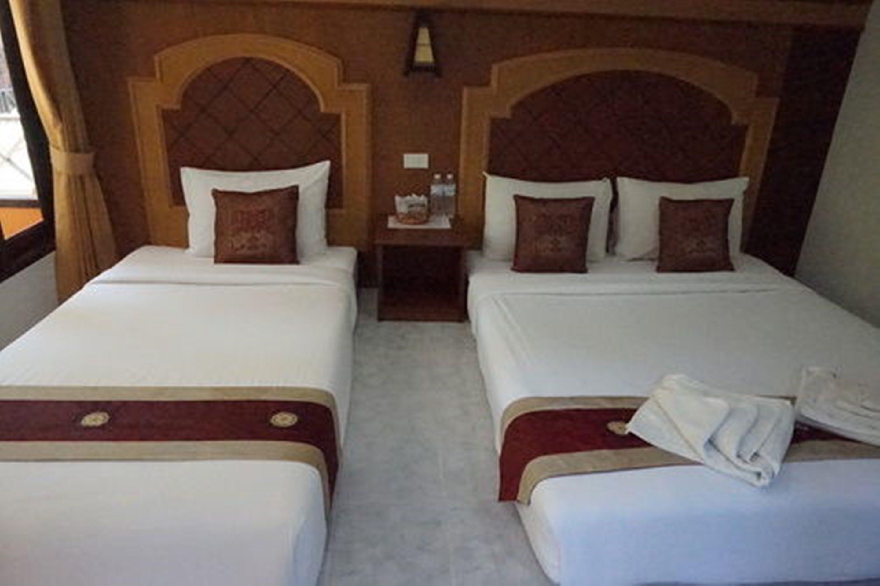 Quarto de hotel com duas camas, cada uma com cabeceira, mesa de cabeceira e janela com cortinas.