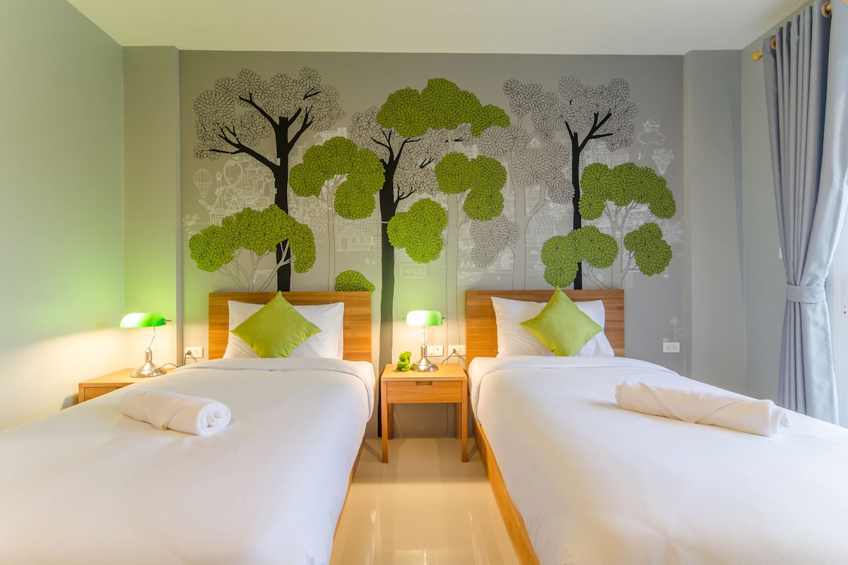 Quarto de hotel com duas camas, almofadas verdes, um mural de árvores na parede e uma janela com cortinas.