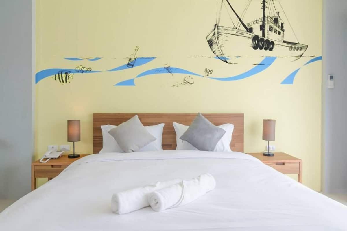 Quarto de hotel com uma cama, duas mesas de cabeceira, um mural de navio na parede e uma toalha enrolada na cama.