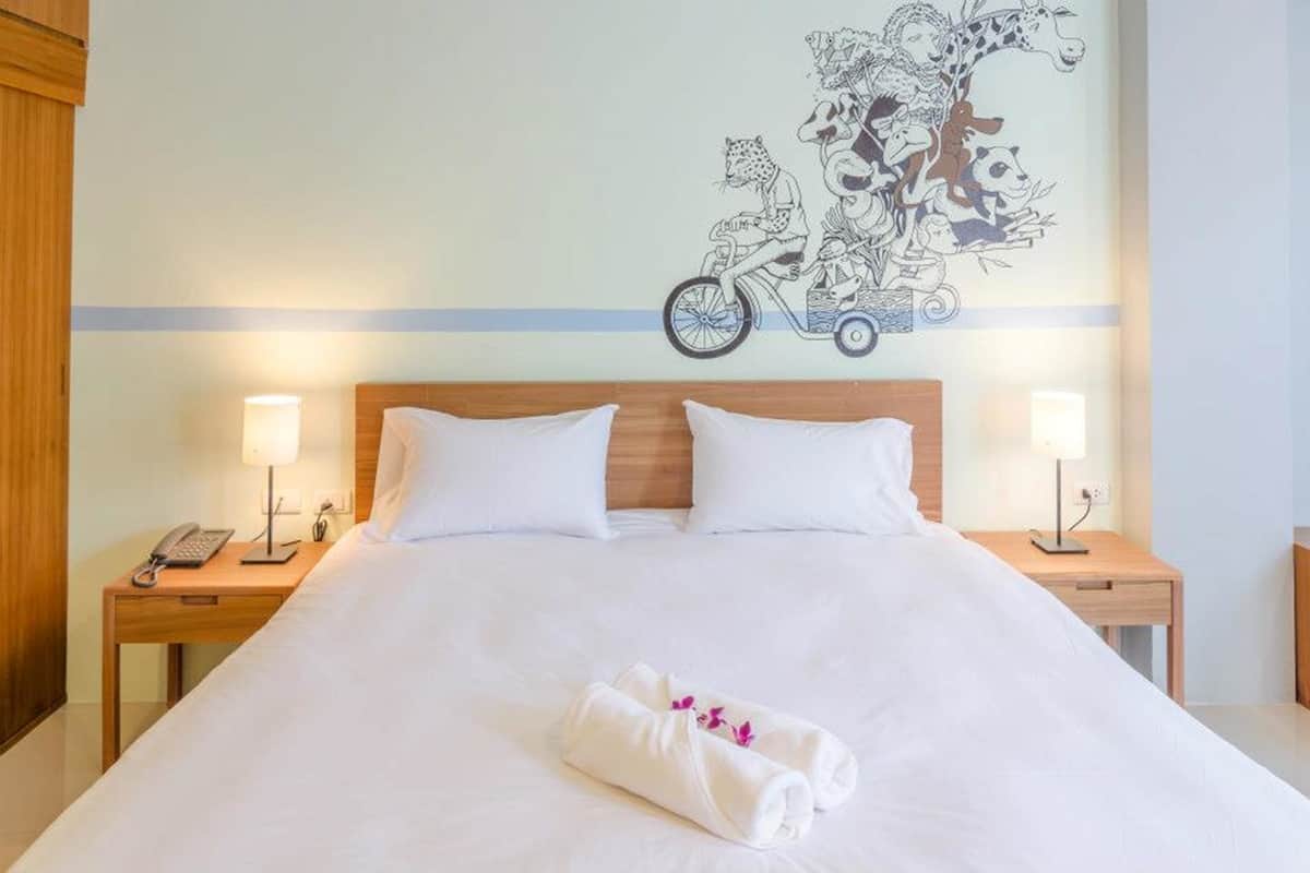 Cama bem arrumada com lençóis brancos, duas mesas de cabeceira com abajures e um mural na parede.