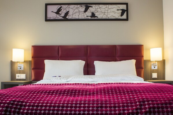 Quarto de hotel com uma cama grande, duas almofadas brancas, cabeceira vermelha e duas luminárias de apoio.