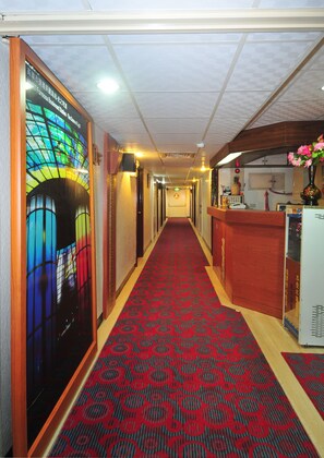 Corredor de hotel com carpete vermelho, painéis de madeira, balcão de recepção e uma janela de vitrais colorida.