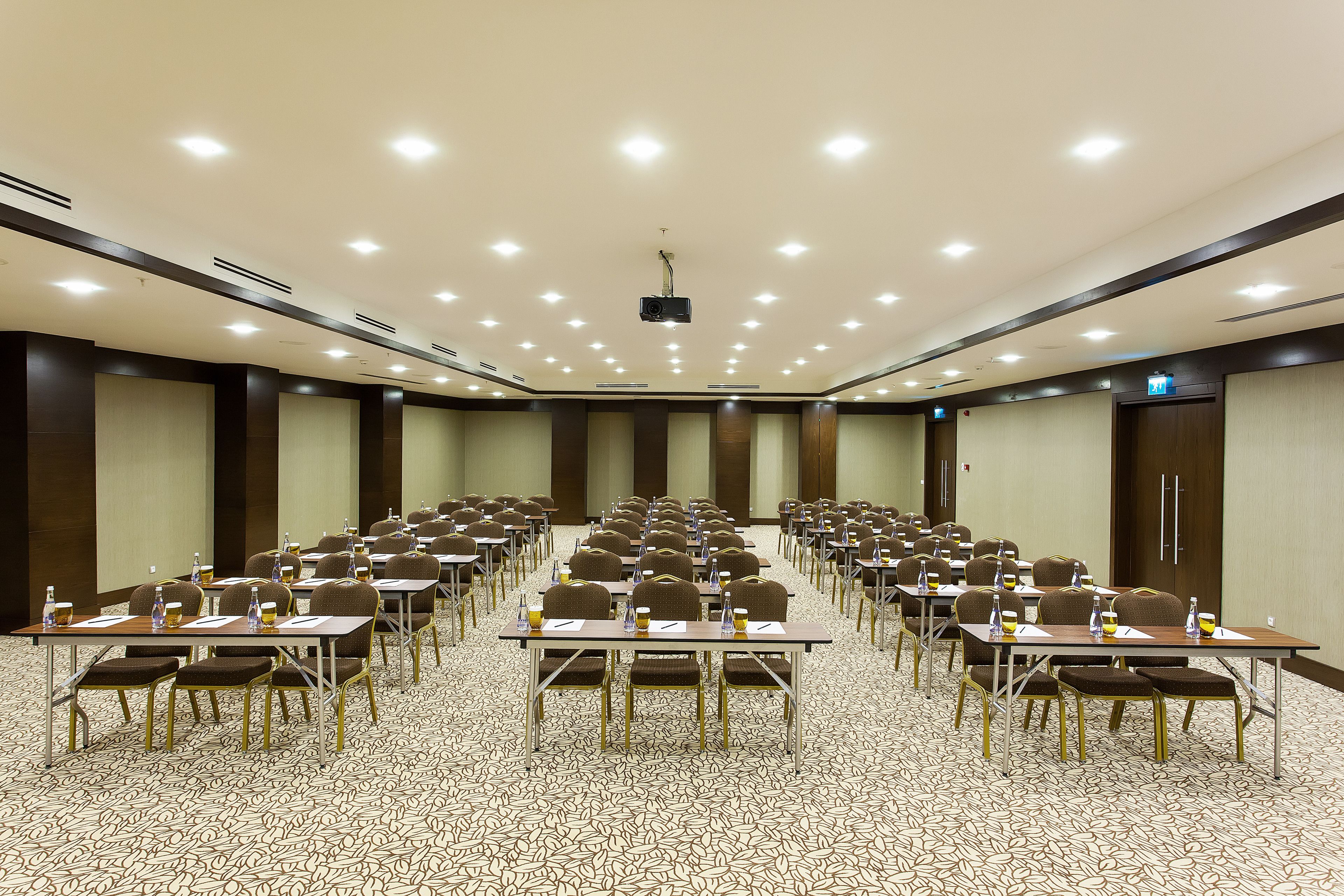 Uma sala de conferências com mesas e cadeiras dispostas em fileiras, equipada com garrafas de água e blocos de anotações.