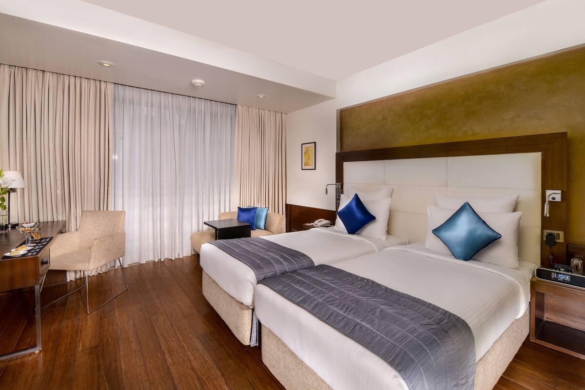 Quarto de hotel com duas camas, uma escrivaninha, uma cadeira, uma mesa de cabeceira e uma janela grande com cortinas.