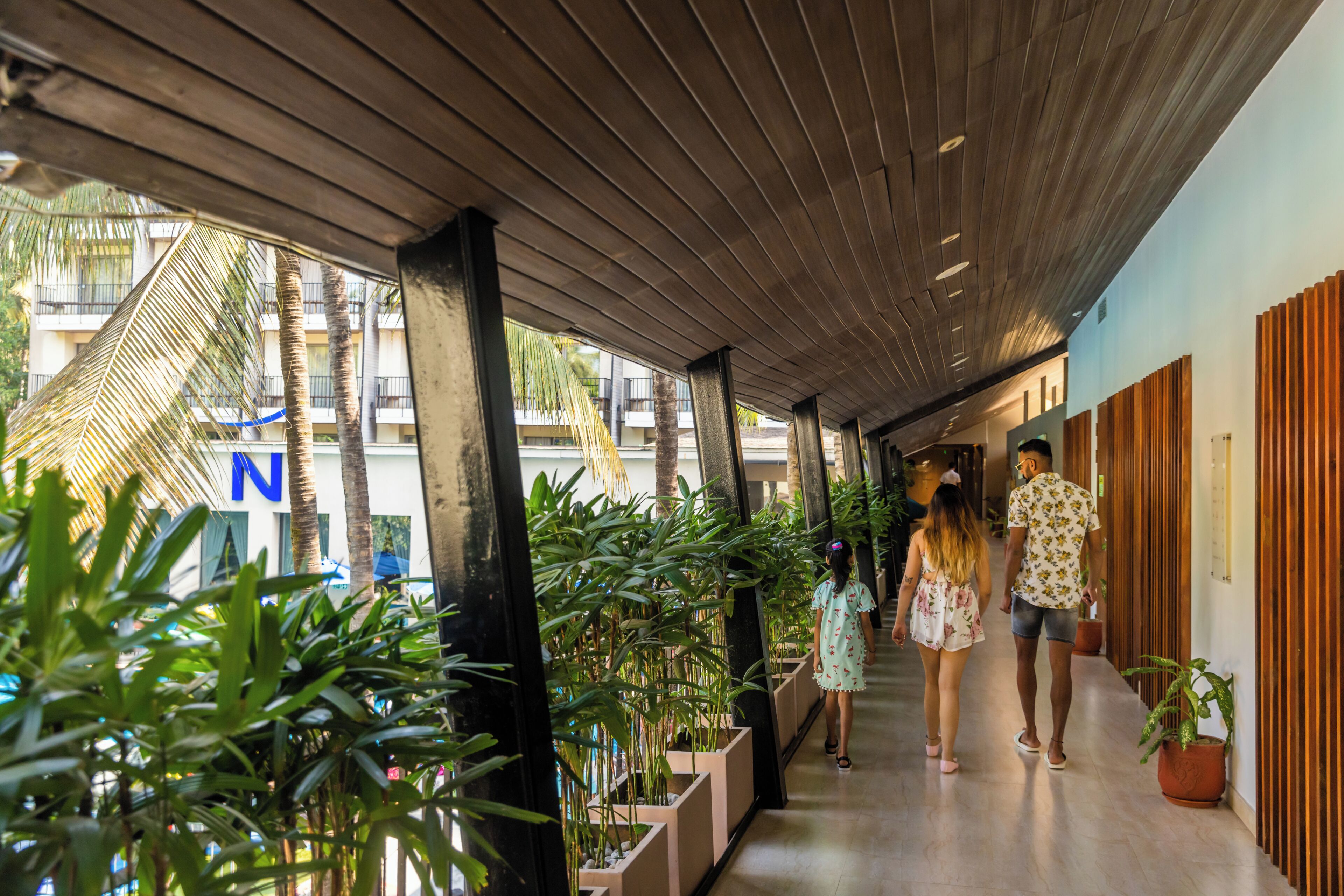 Uma galeria coberta com vigas de madeira, plantas em vasos e vista de edifícios ao fundo.