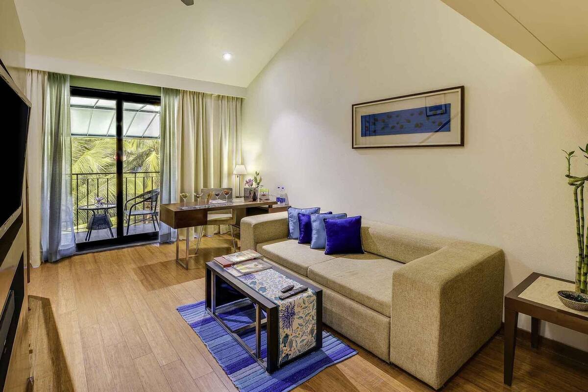 Uma sala de estar com sofá, mesa de centro e área de jantar com mesa e cadeiras. Há um ventilador de teto, um quadro na parede e uma planta sobre uma mesinha lateral.