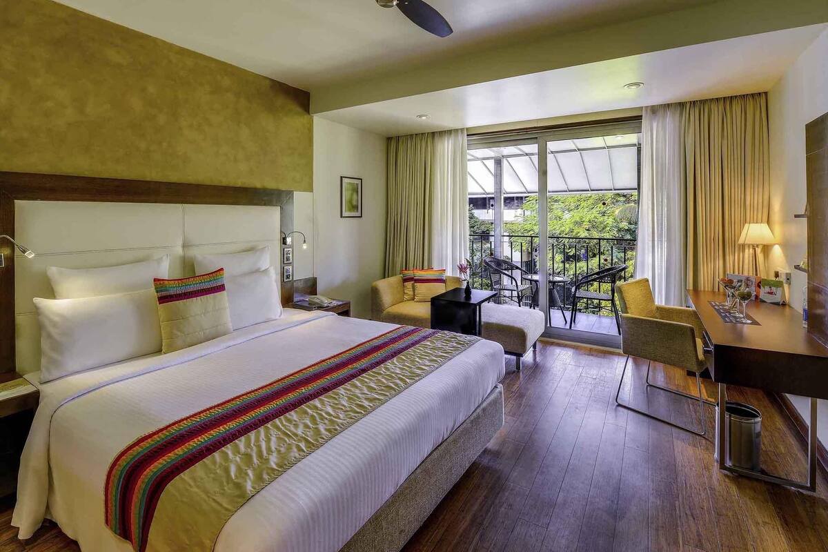 Quarto de hotel com uma cama grande, uma escrivaninha, uma cadeira e varanda com vista para o verde.