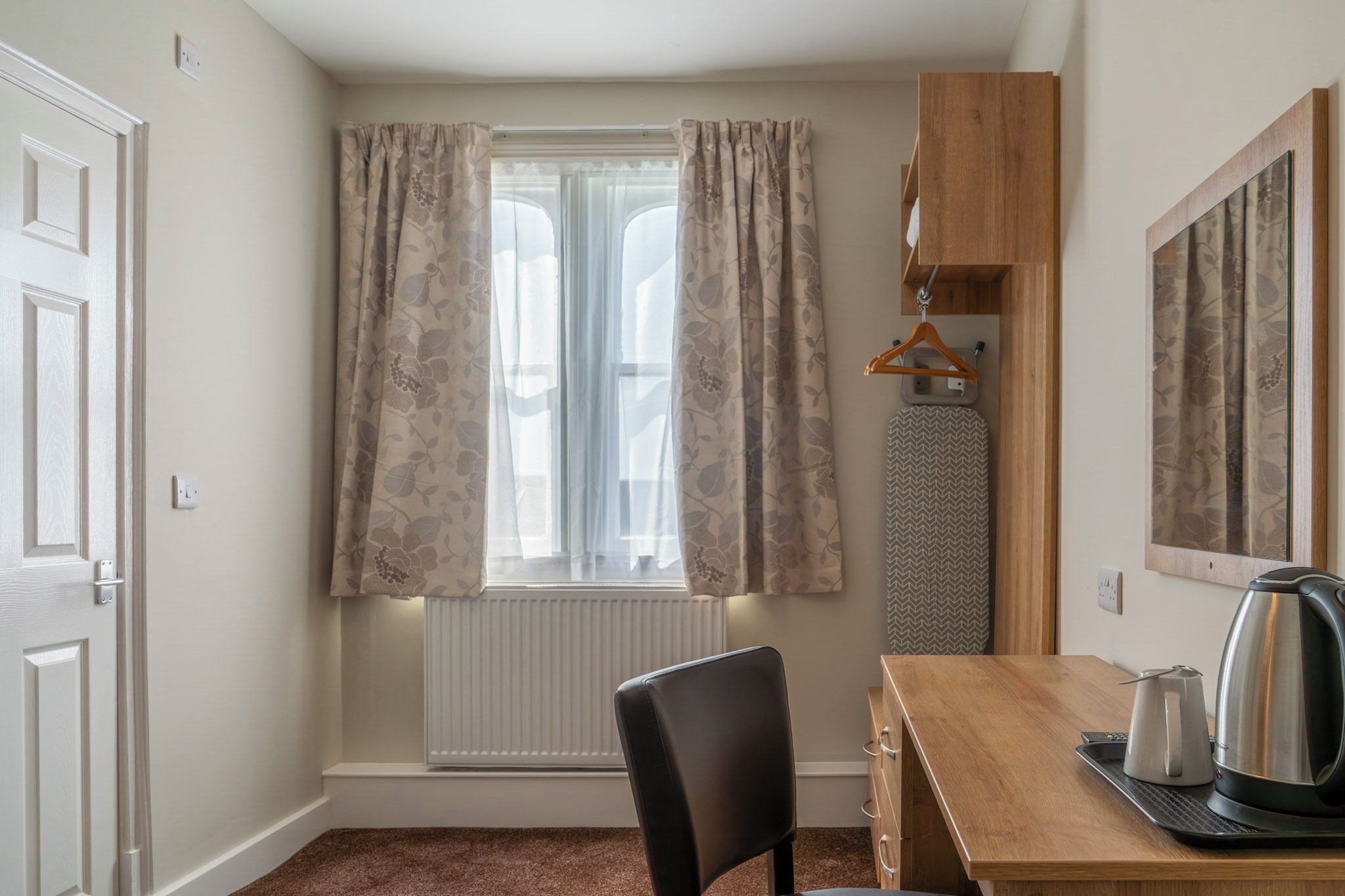 Quarto com escrivaninha de madeira, cadeira, radiador, janela com cortinas, cabideiro e chaleira.
