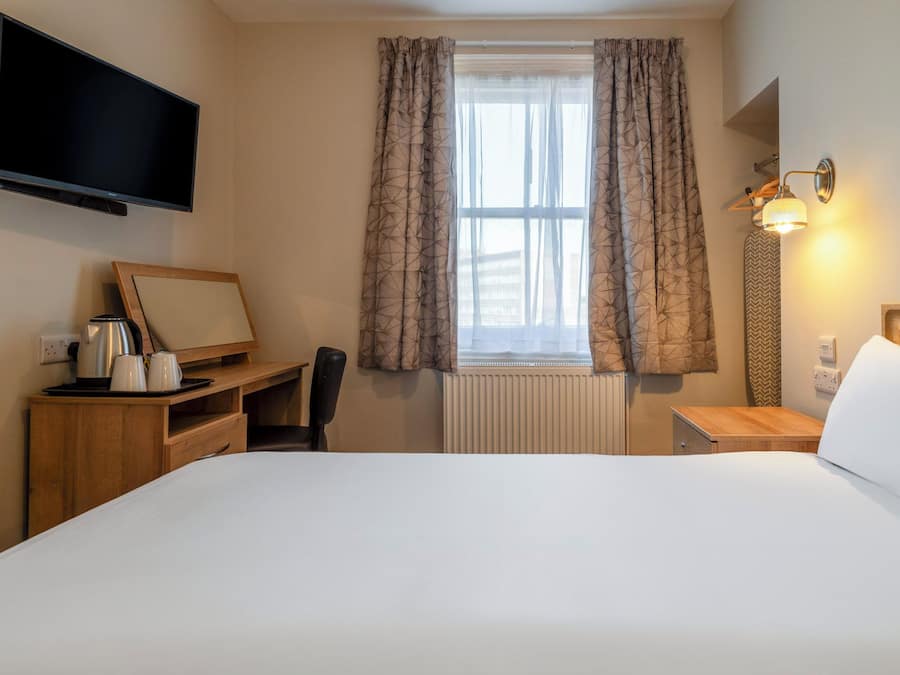 Quarto de hotel com uma cama, uma escrivaninha com cadeira, televisão, janela com cortinas e abajur.