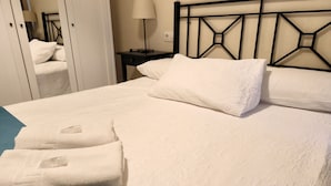 Cama bem arrumada com lençóis brancos e um toque de azul, uma luminária de cabeceira e um espelho refletindo o quarto.