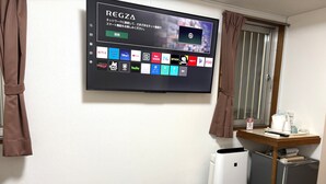 Quarto de hotel com TV fixada na parede exibindo diversos serviços de streaming, uma escrivaninha com cafeteira, lixeira e um refrigerador compacto.