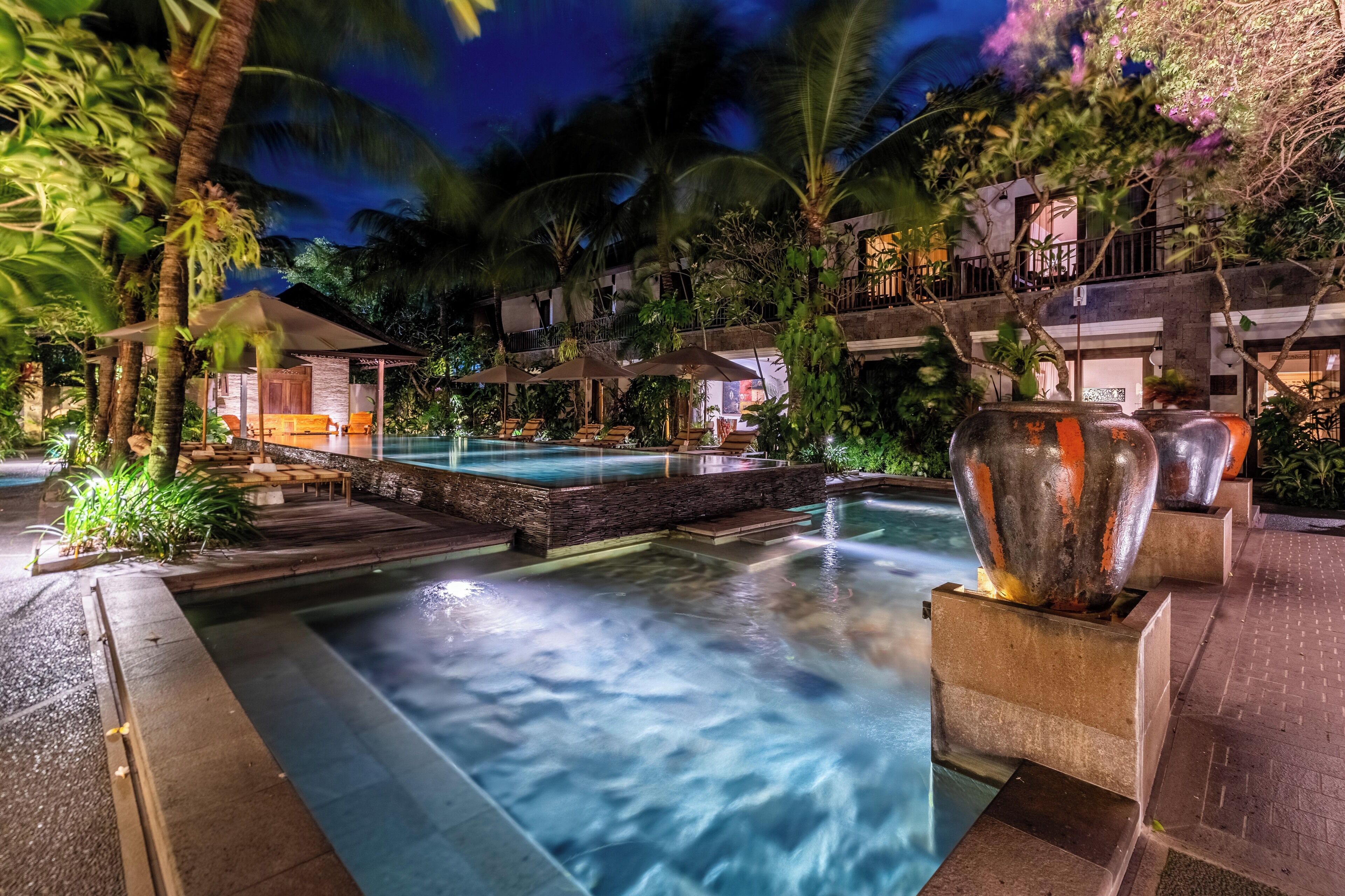 Área de piscina noturna em resort, com hidromassagem, espreguiçadeiras e um caminho de pedra.