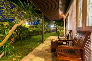 Área de varanda externa com bancos de madeira e um caminho de concreto, cercada por vegetação exuberante e uma estrutura de pérgola de madeira.