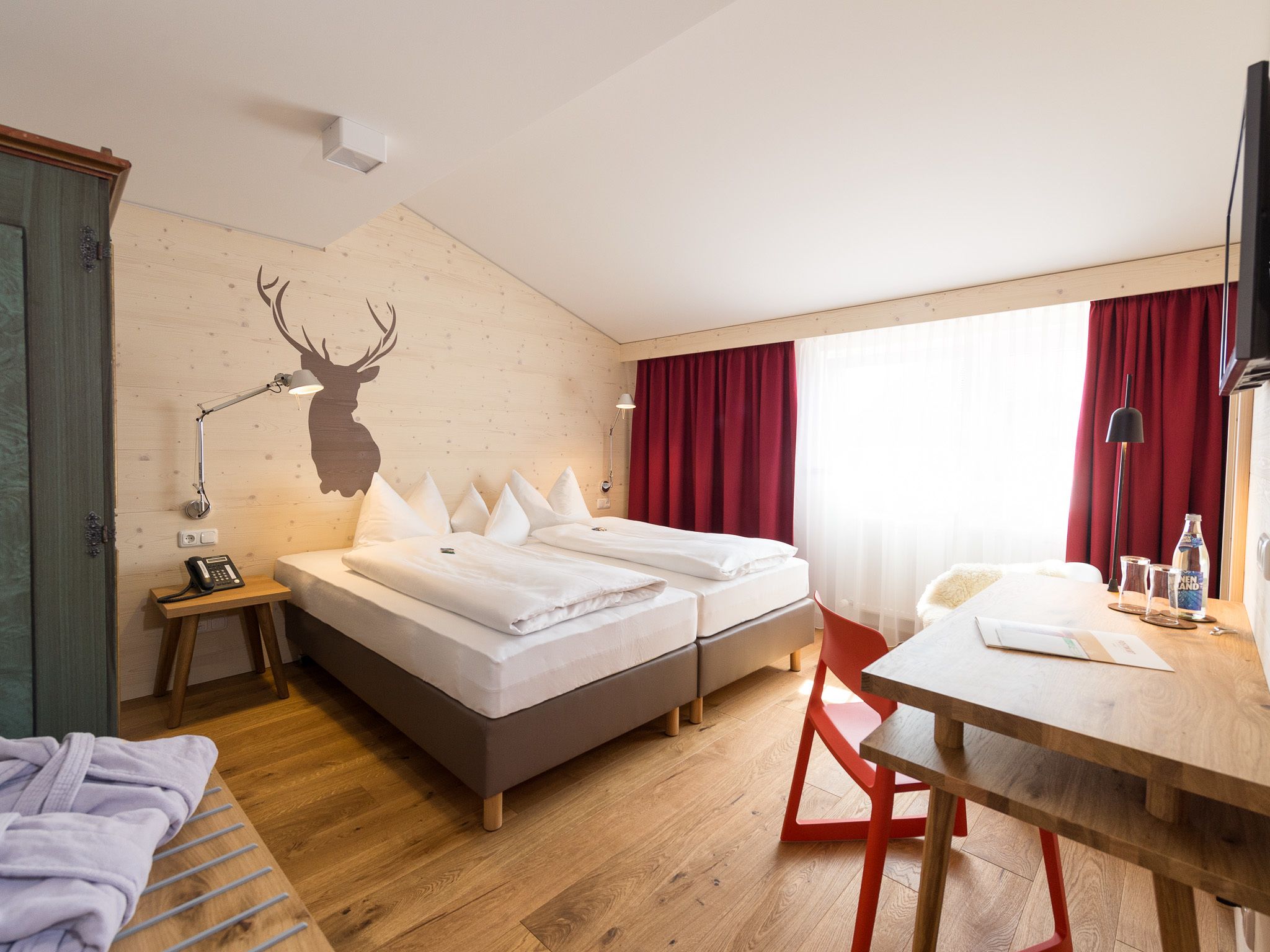 Quarto de hotel com piso de madeira, uma cama com roupa de cama branca, uma escrivaninha com cadeira, cortina vermelha e uma luminária fixada na parede.
