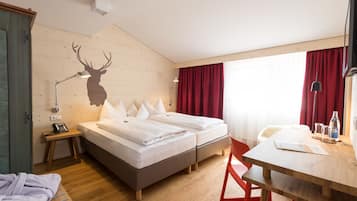 Quarto de hotel com piso de madeira, uma cama com roupa de cama branca, uma escrivaninha com cadeira, cortina vermelha e uma luminária fixada na parede.