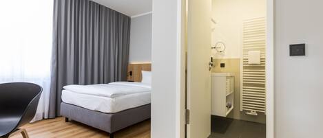 Quarto de hotel moderno com cama, poltrona, banheiro com vaso sanitário e pia, e vista para o exterior através de uma janela com cortinas.