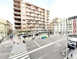 Um edifício residencial com várias andares e varandas, situado numa área urbana com passadeira e paragem de autocarro.