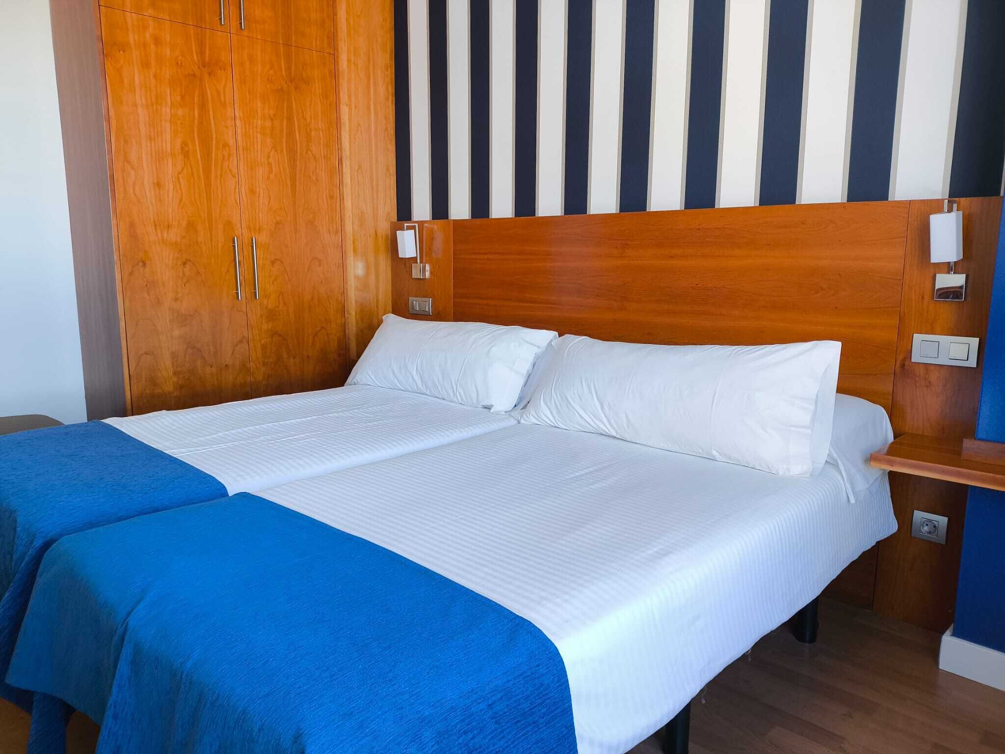 Cama de casal com roupa de cama branca e mantas azuis, cabeceira de madeira e papel de parede listrado.