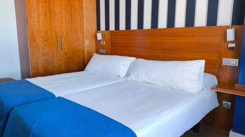 Cama de casal com roupa de cama branca e mantas azuis, cabeceira de madeira e papel de parede listrado.