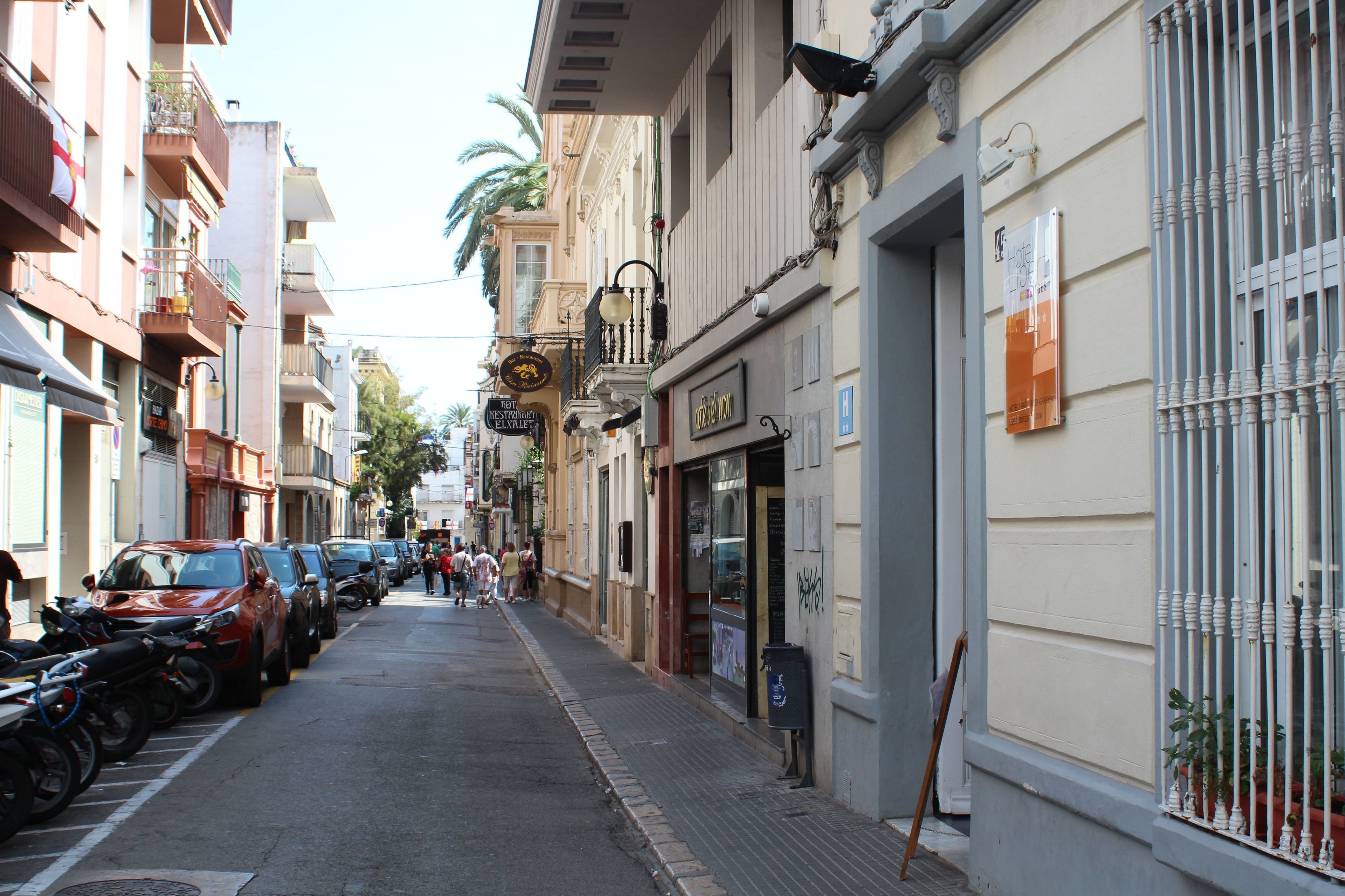 Rua estreita flanqueada por carros estacionados e edifícios com varandas.