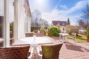 Uma área de pátio ao ar livre com mesa e cadeiras, um hedge e uma casa ao fundo.