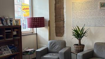 Uma sala de estar com dois poltronas cinzas, uma estante, um abajur vermelho, um vaso de plantas e uma parede com um painel de texto grande.