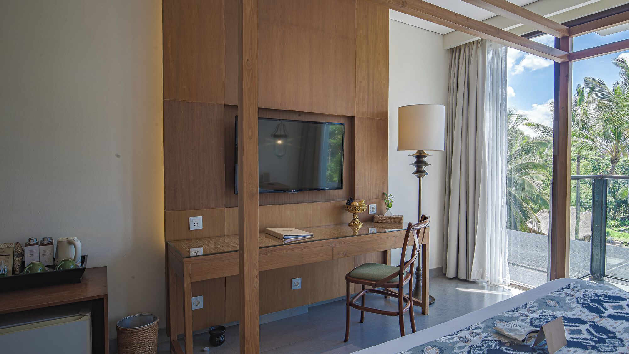 Um quarto com escrivaninha de madeira, televisão, luminária, cadeira e vista para palmeiras pela janela.