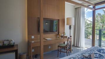 Um quarto com escrivaninha de madeira, televisão, luminária, cadeira e vista para palmeiras pela janela.