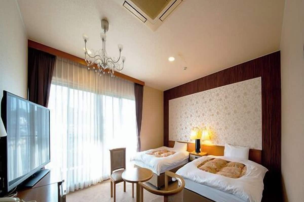 Quarto de hotel com duas camas, televisão, candelabro e uma janela grande com cortinas.